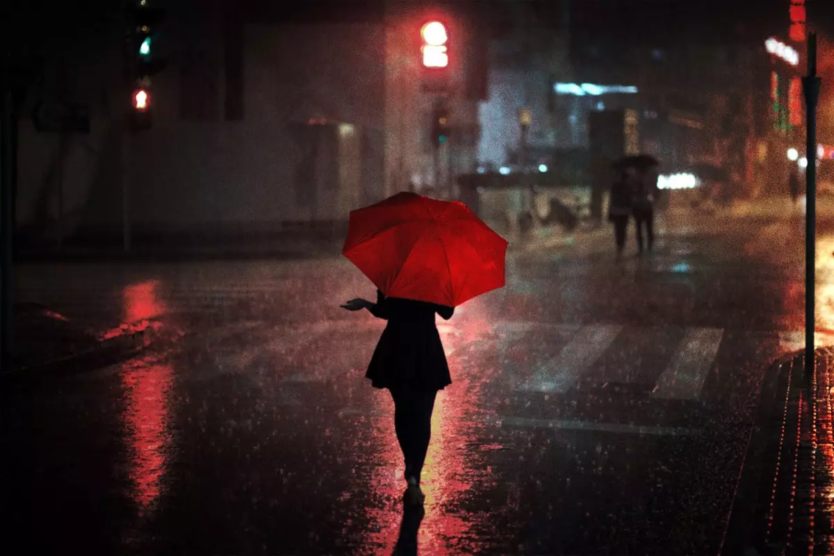 O femeie din Shanghai, China apare în siluetă în timp ce traversează o stradă ploioasă noaptea. Își întinde mâna sub o umbrelă roșie