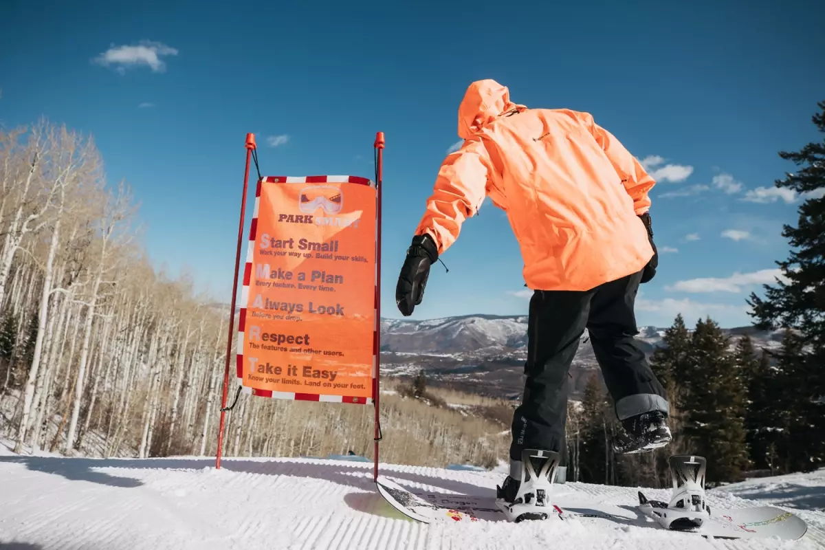 Profesionalus snieglentininkas Brockas Crouchas važiuoja Burton Step On apkaustais be petnešėlių. Čia jis pavaizduotas oranžine žiemine striuke ant snieguoto kalno viršūnėje Aspene, Kolorado valstijoje, šalia oranžinio ženklo.