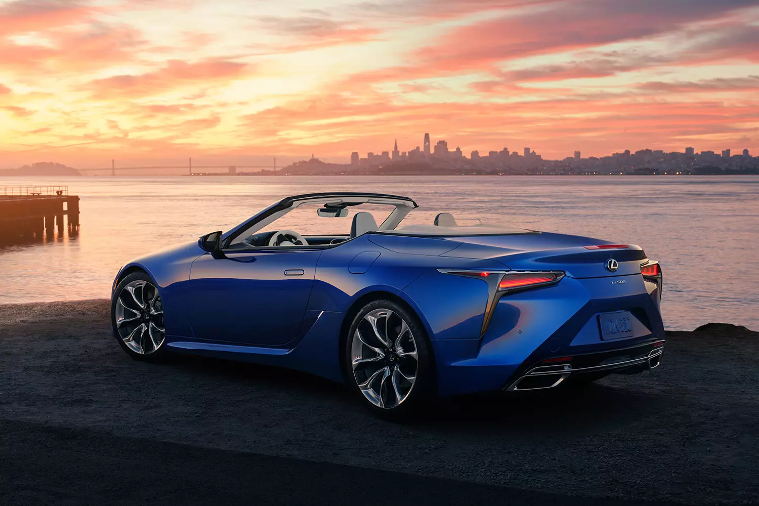 Το πίσω άκρο ενός μπλε Το πίσω άκρο του Lexus LC 500 Cabrio 2021 με μια πόλη και το ηλιοβασίλεμα στο βάθος. Δοκιμάσαμε το grand tourer και το προτείνουμε ολόψυχα.