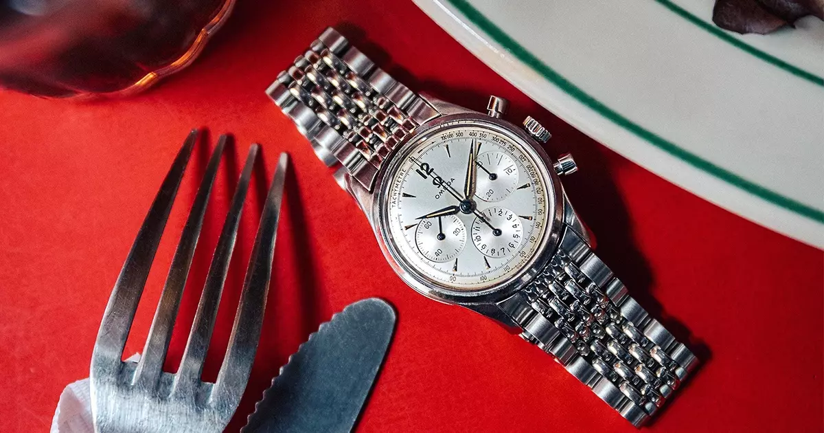 Senovinis Omega Seamaster Chronograph sėdi ant raudonos staltiesės ant valgomojo stalo šalia šakutės, peilio ir lėkštės. Senovinis laikrodis šiuo metu parduodamas per J.Crew.