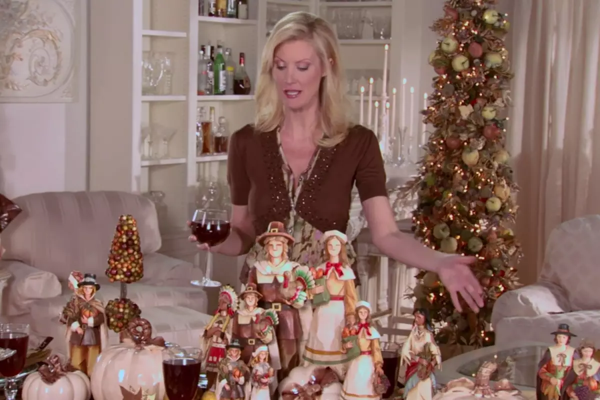 Ѕвездата на Food Network, Сандра Ли држи чаша црвено вино и гестикулира кон масата на Денот на благодарноста со фигурини на аџии во оваа слика од екранот од Полу-домашно готвење со Сандра Ли