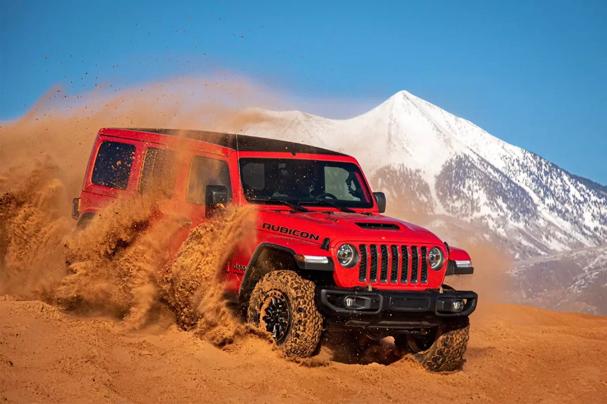 ລົດ Jeep Wrangler Rubicon 392 ສີແດງ, ເປັນ Wrangler ທໍາອິດທີ່ເຄີຍມີເຄື່ອງຈັກ V8, ປັ່ນປ່ວນດິນຊາຍຢູ່ທາງຫນ້າຂອງພູເຂົາທີ່ມີຫິມະປົກຄຸມ.