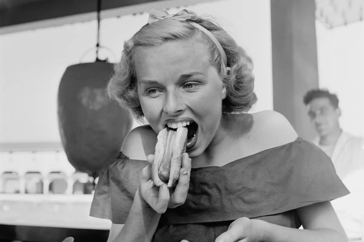 La foto en blanco y negro muestra a una mujer joven comiendo un perrito caliente en Palisades Park, Nueva Jersey, 1949.