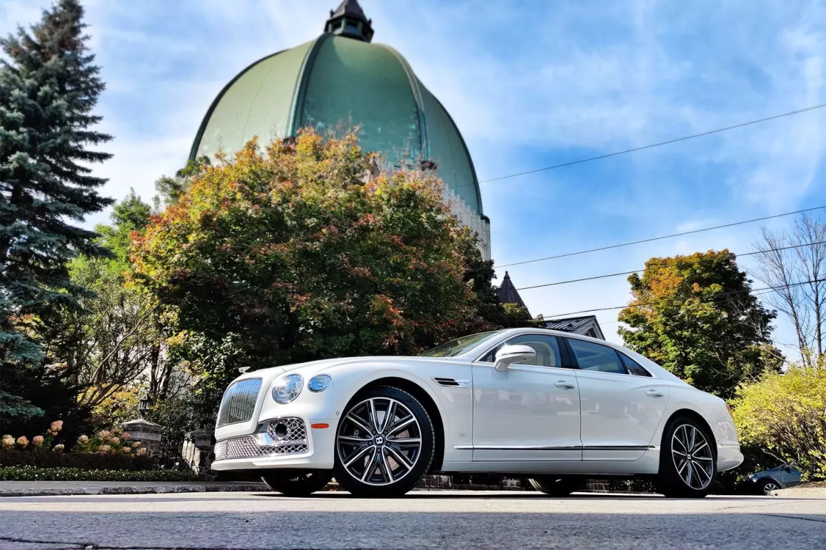 Седанот Bentley Flying Spur од 2022 година во бело седи пред дрвја и зграда со бакарна купола