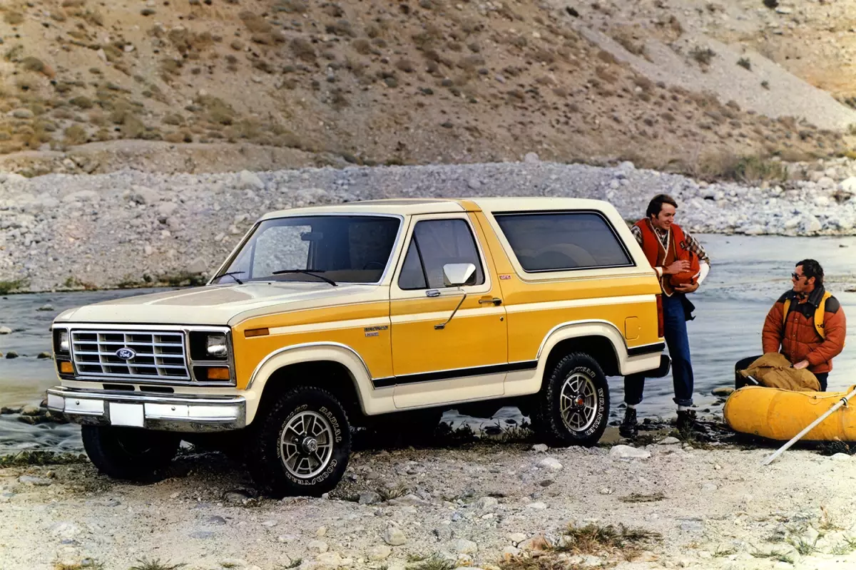 在一張復古照片中，一輛白色和黃色的 1982 年福特 Bronco SUV 坐在岩石和河流中，旁邊是兩個戶外運動者