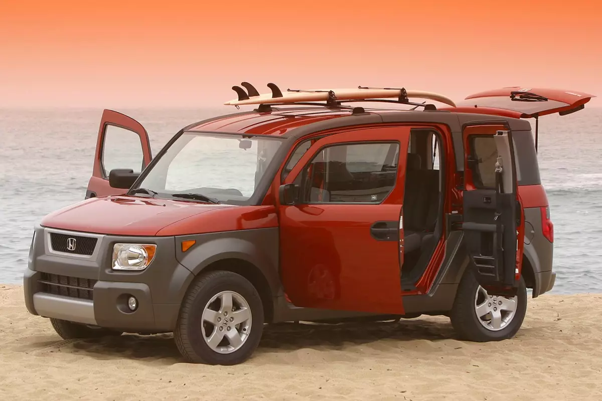 Un Honda Element laranxa e marrón con todas as portas abertas e unha táboa de surf enriba sentada na praia ao pór do sol