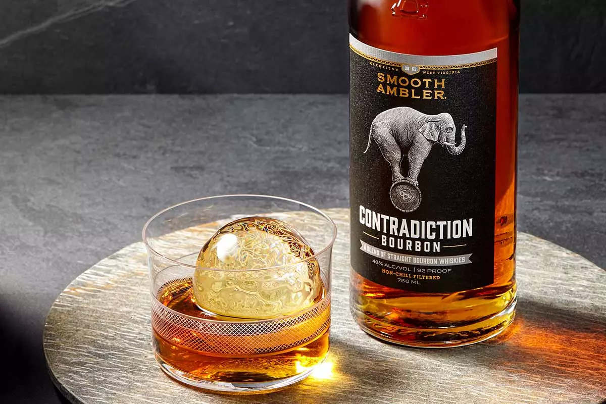 Una botella de bourbon Contradiction y un vaso de whisky con una bola de hielo